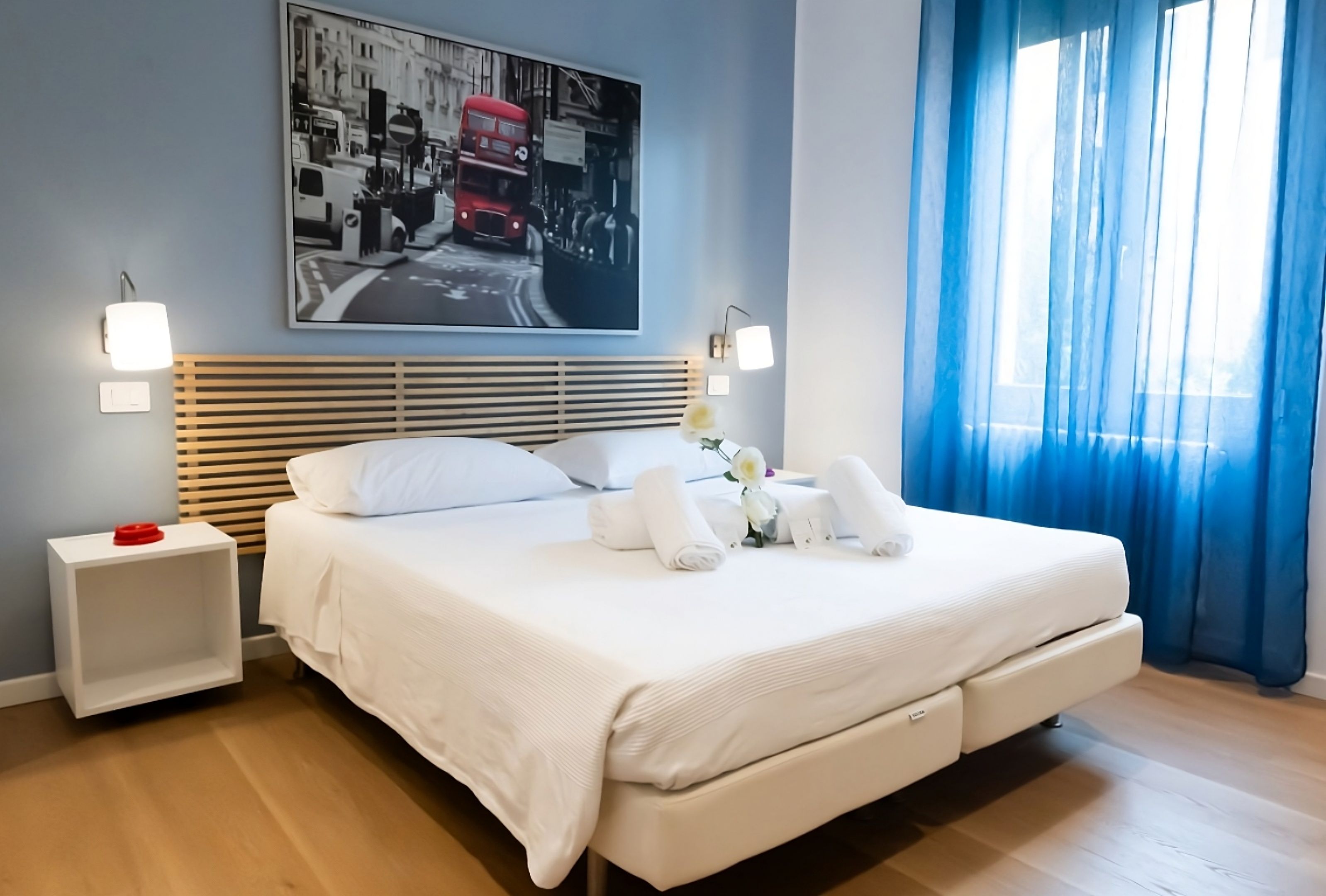 Uma cama bem arrumada com lençóis brancos, uma cabeceira com ripas horizontais, uma mesa de cabeceira com um objeto vermelho, um quadro na parede de um ônibus vermelho e uma janela com cortinas azuis.