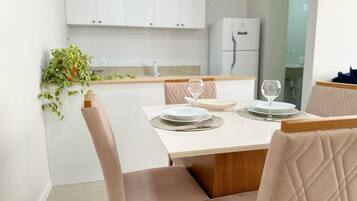 Uma área de refeições com uma mesa branca posta para quatro pessoas, rodeada por cadeiras estofadas com encosto acolchoado. É visível uma cozinha com armários brancos e uma geladeira ao fundo.