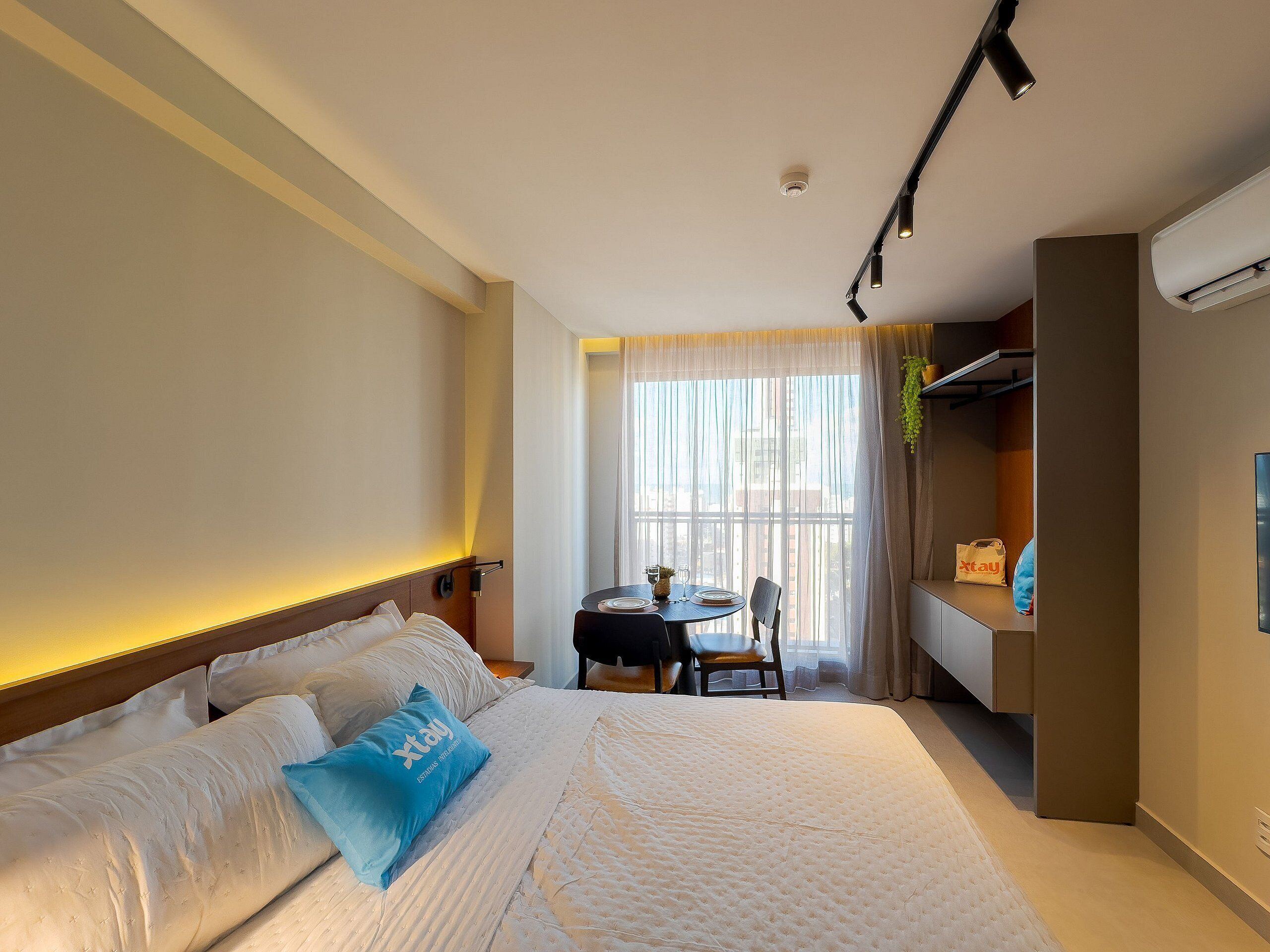 Quarto de hotel com cama, escrivaninha e cadeira. Há uma janela com cortinas e um aparelho de ar condicionado na parede.