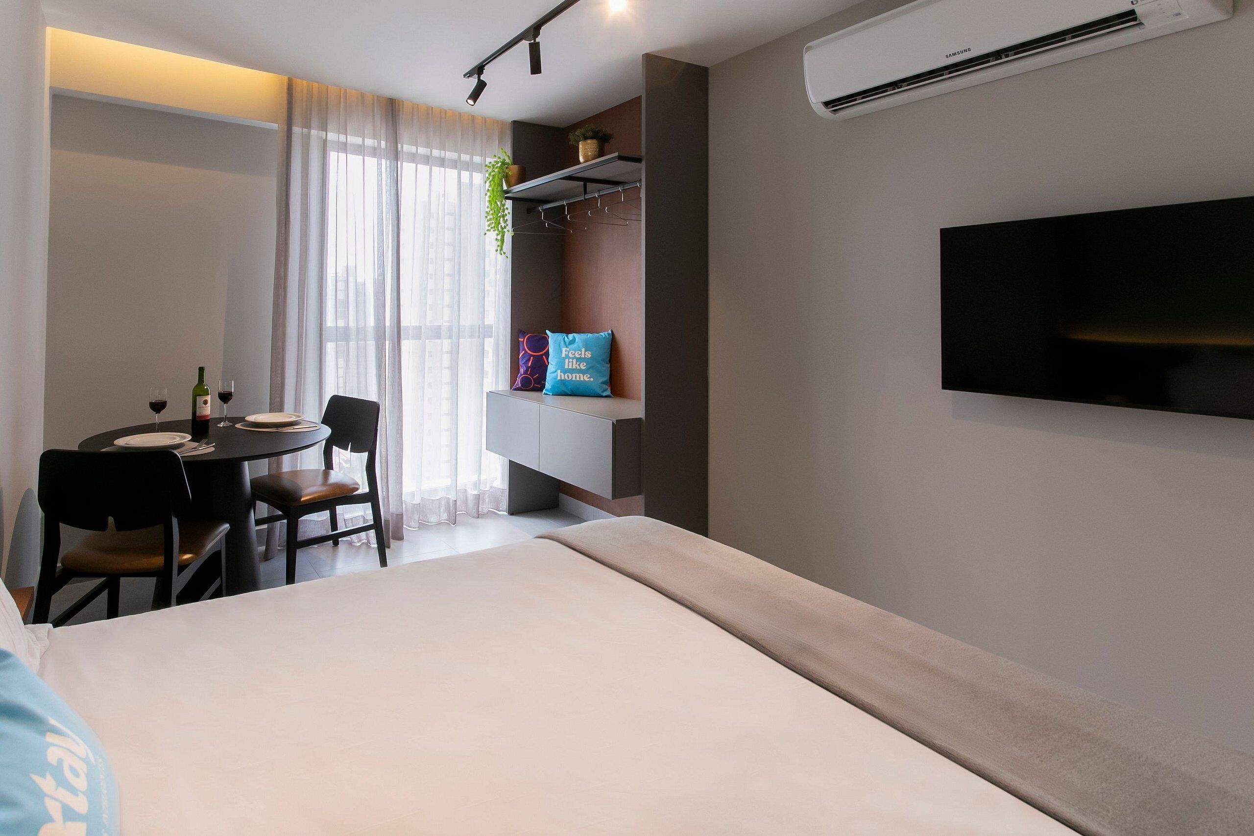 Quarto de hotel moderno com uma cama, área de refeições com mesa e cadeiras, TV fixada na parede e aparelho de ar condicionado.