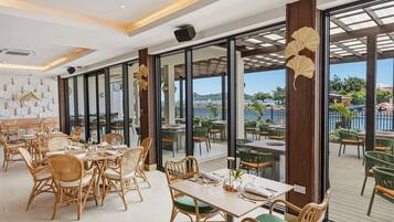 Restaurante com mesas dentro e fora, grandes janelas e vista para o mar.