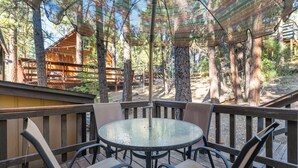 Um deck de madeira com uma mesa de vidro redonda e cadeiras, rodeado por árvores e uma cabana de madeira ao fundo.