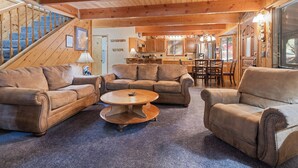 Interior acolhedor de uma cabana com escada de madeira, dois sofás, uma mesa de centro e um espaço de jantar com mesa e cadeiras.