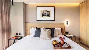 Quarto de hotel bem arrumado com uma cama grande, uma mesa de cabeceira com um livro, um quadro na parede e uma mesinha de noite com uma luminária.