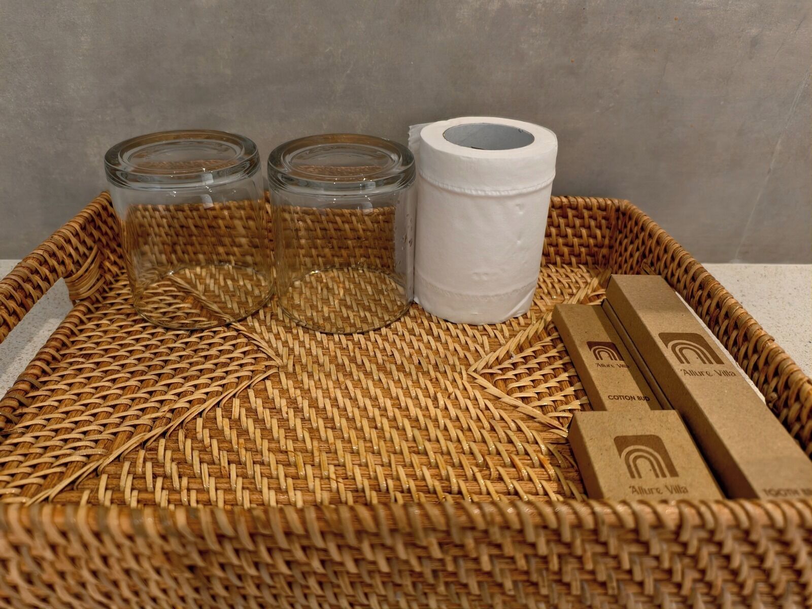 Uma cesta de vime com três frascos de vidro, um rolo de papel higiênico e duas caixas de cartão com a marca.