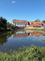 Um edifício com telhado de telhas vermelhas, um lago à frente e vegetação ao redor.