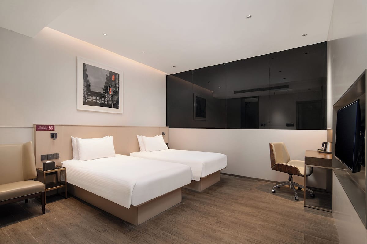 Quarto de hotel com duas camas, uma escrivaninha, uma cadeira, uma televisão e um quadro na parede.