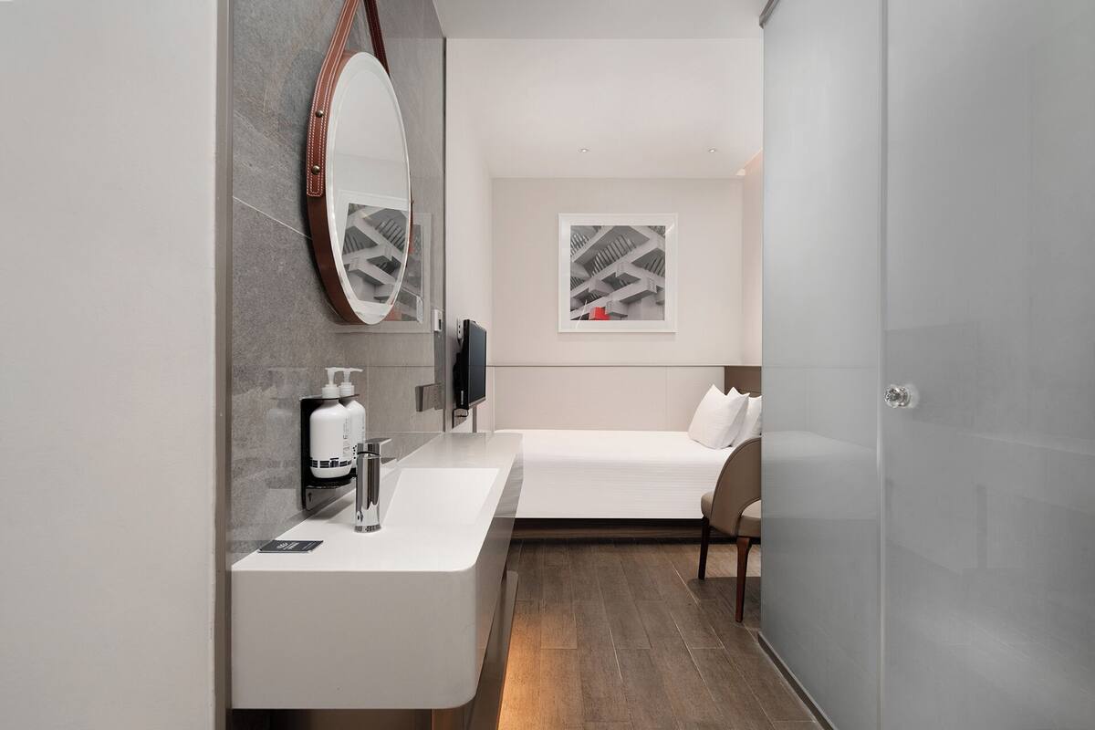 Um banheiro de hotel moderno, com um espelho grande, pia com artigos de higiene, cama e uma moldura com uma fotografia na parede.