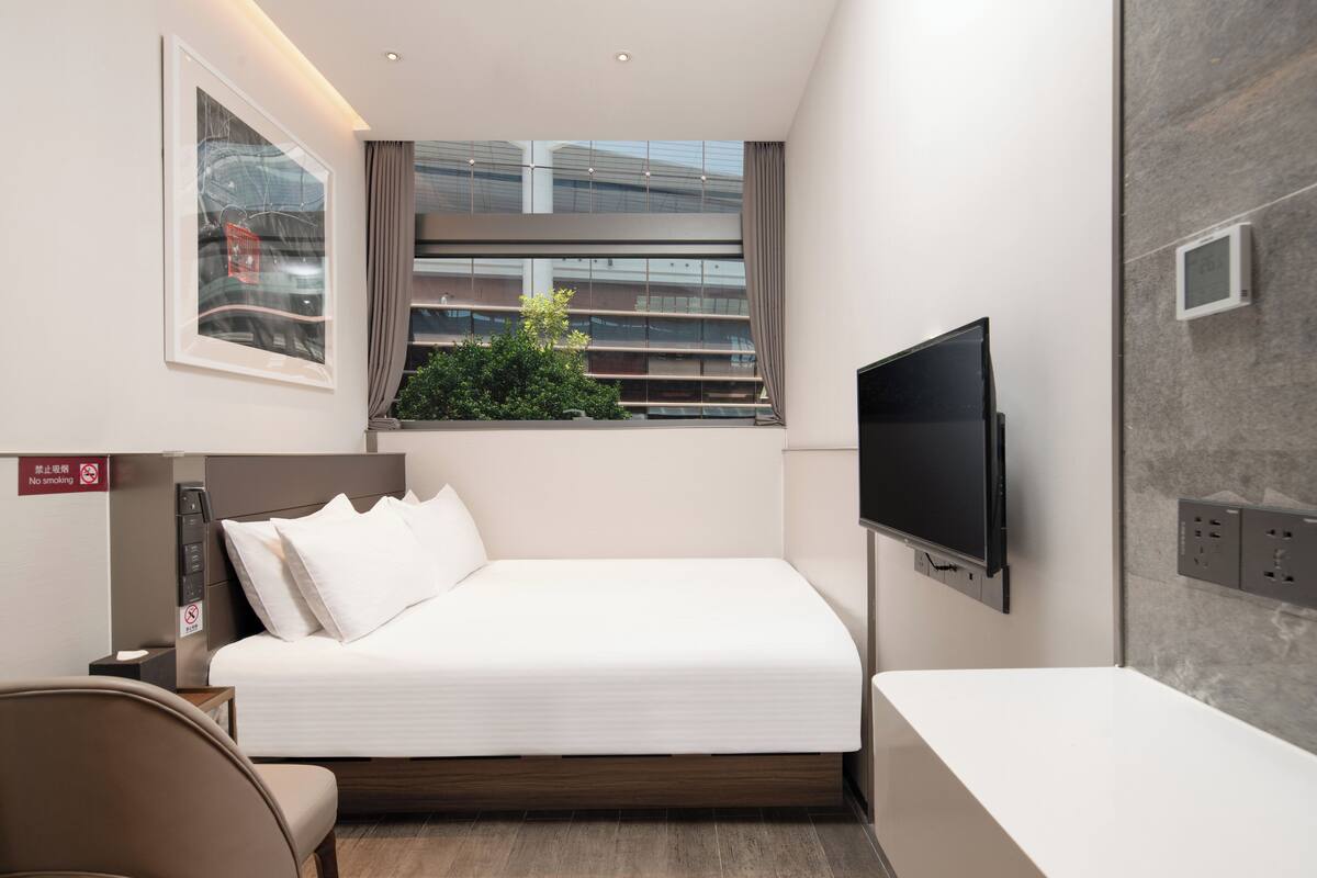 Quarto de hotel moderno com cama, televisão, escrivaninha e janela com vista para prédios.