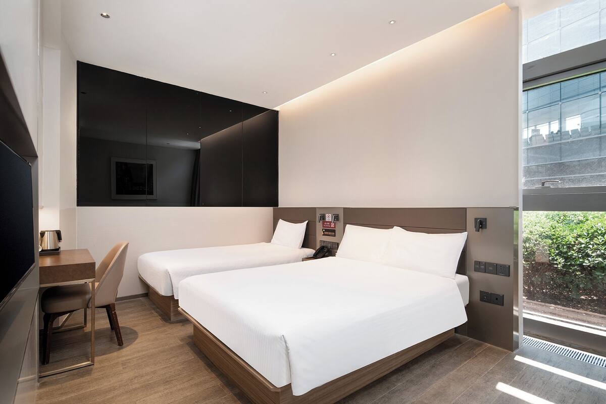 Quarto de hotel moderno com duas camas, uma mesa e uma grande janela com vista para o verde.