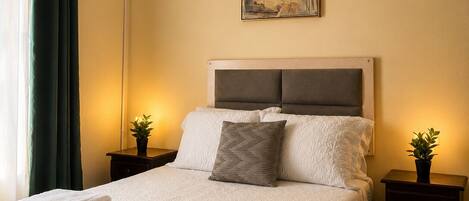 Cama bem arrumada com cabeceira, duas mesas de cabeceira com abajures, um quadro na parede e uma janela com cortinas.
