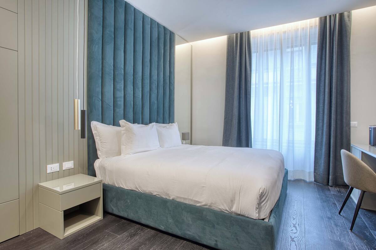 Quarto de hotel moderno com uma cama grande, uma mesinha de cabeceira, uma escrivaninha e uma cadeira.