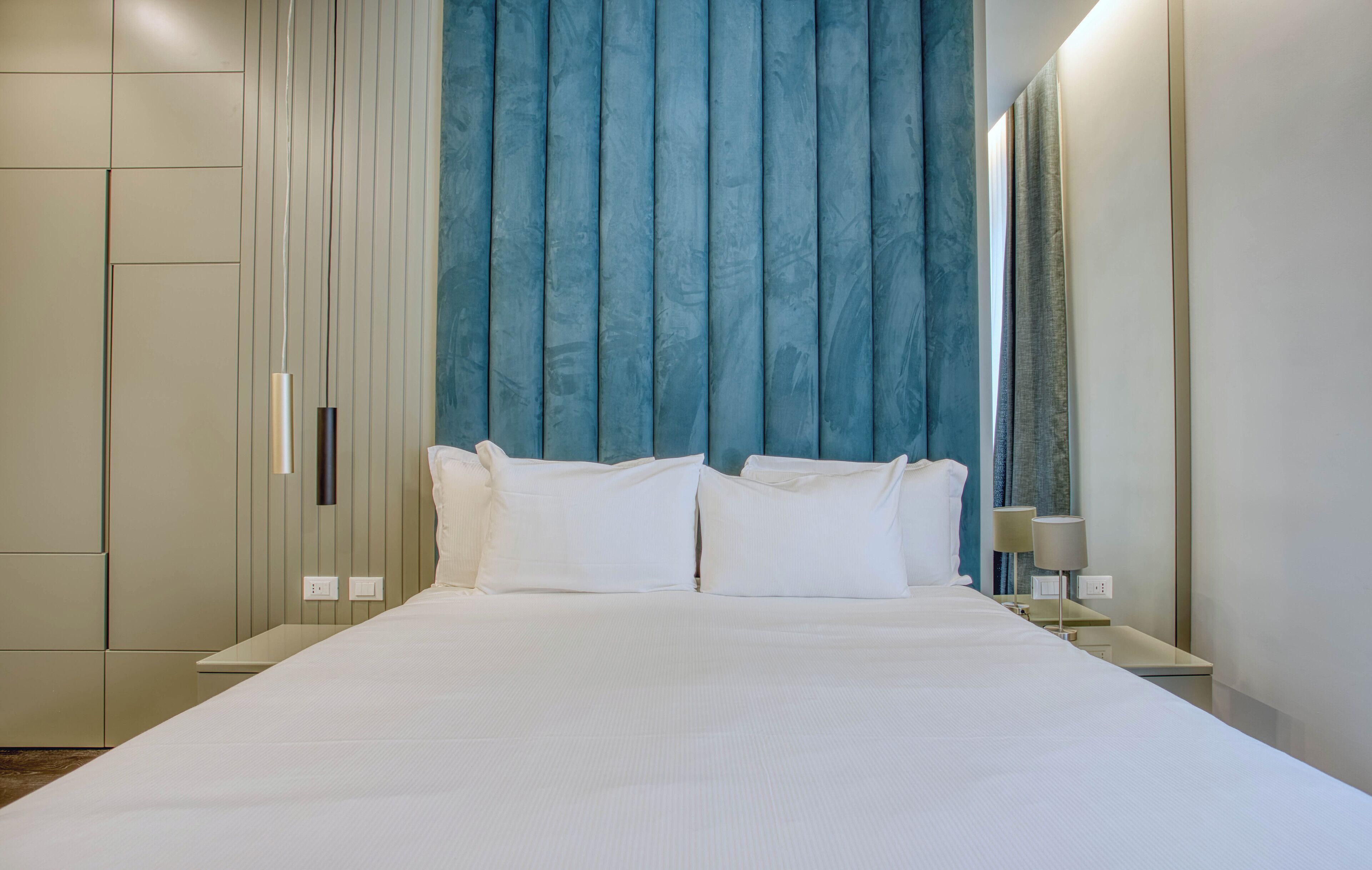 Quarto de hotel moderno com uma cama grande, um guarda-roupa, uma mesa de cabeceira com abajur e uma parede azul de destaque.