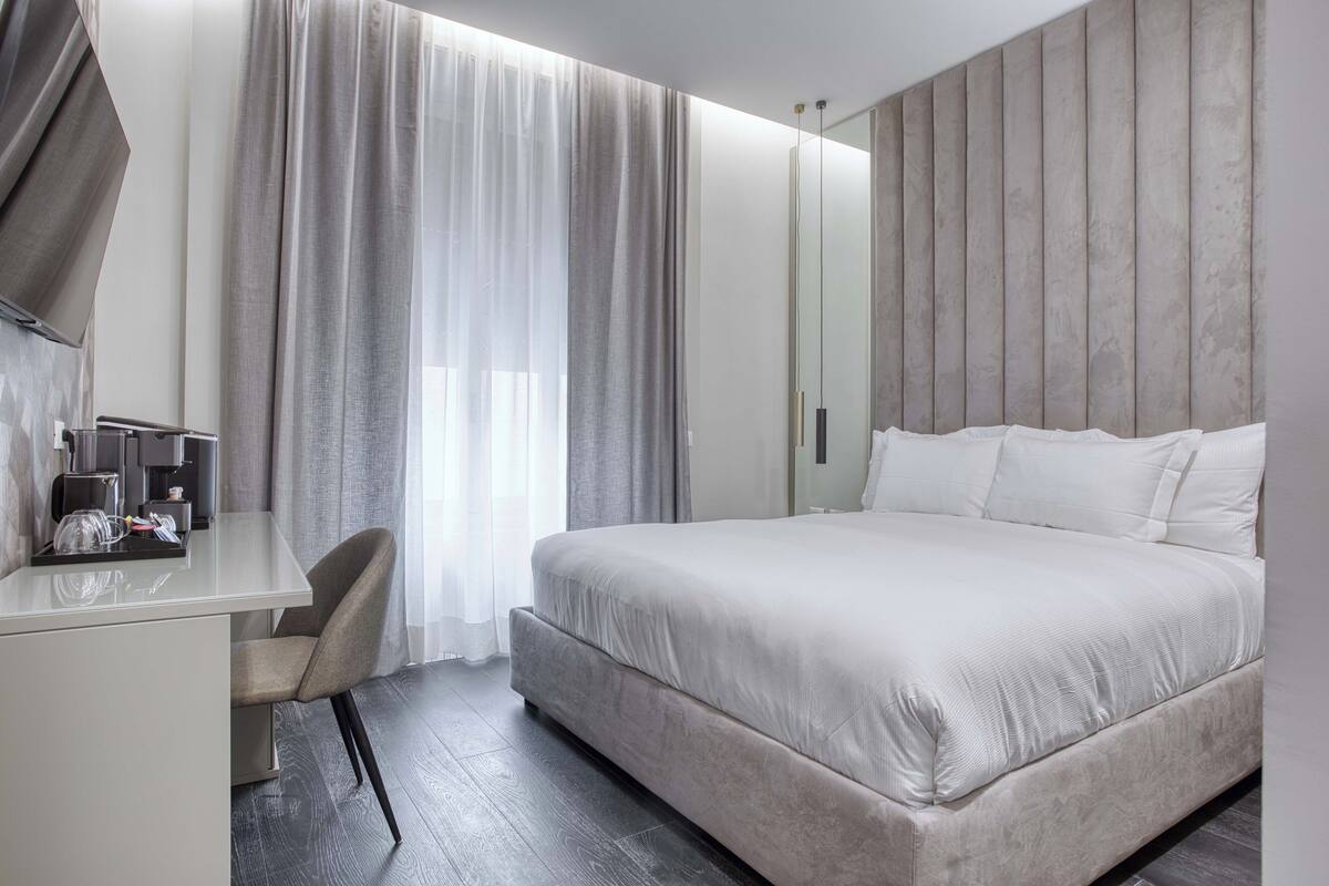 Quarto de hotel moderno com uma cama grande, uma escrivaninha e uma cadeira. Há uma janela com cortinas e um espelho na parede.