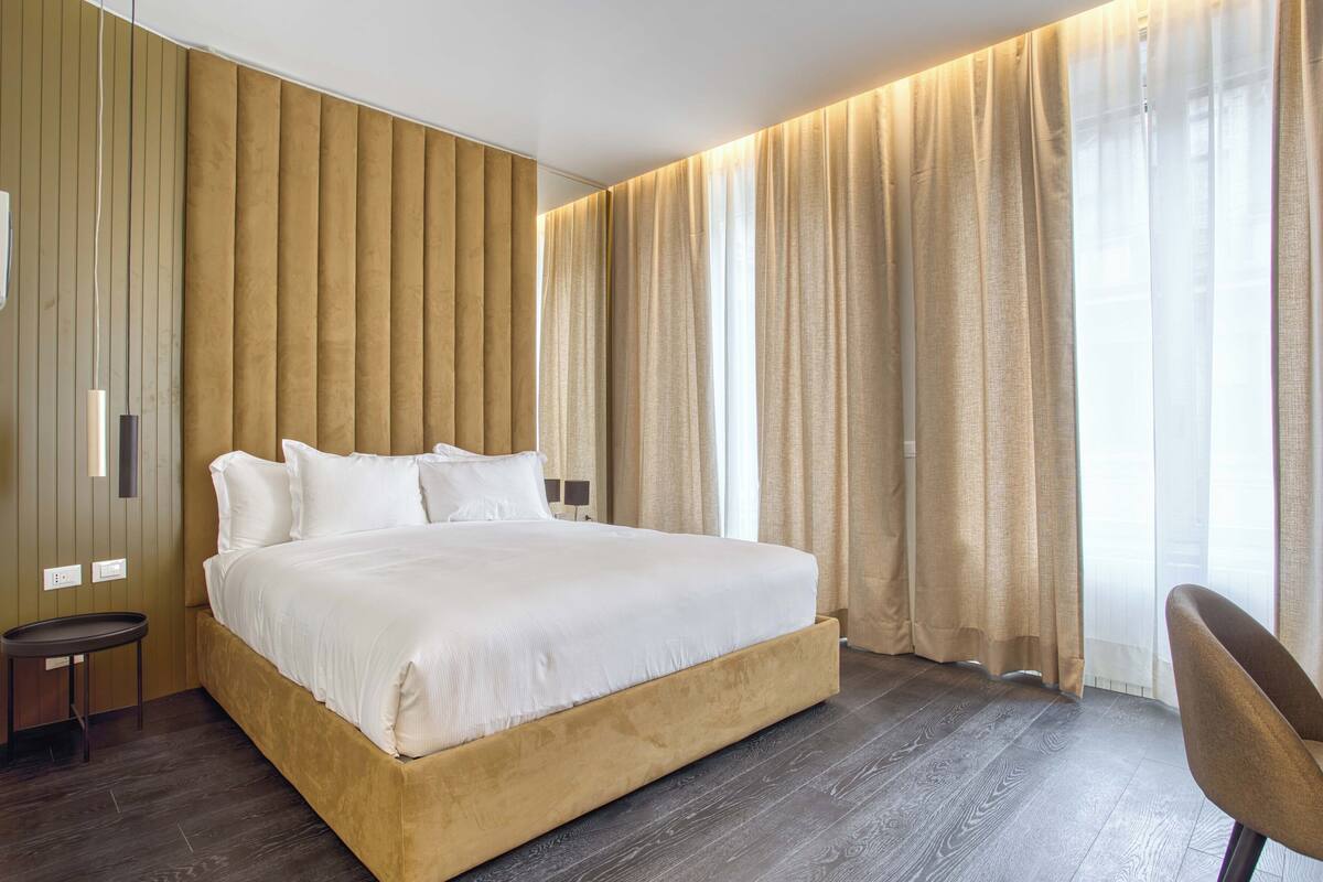 Quarto de hotel moderno com uma cama grande, cabeceira de madeira e vista para a cidade através de cortinas translúcidas.