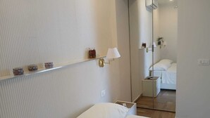 Quarto de hotel com cama, mesa de cabeceira, luminária, prateleira com objetos decorativos e um espelho refletindo o ambiente.