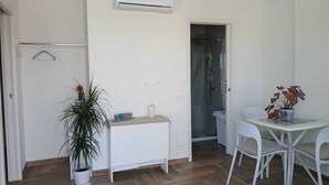 Quarto moderno e bem iluminado, com um ar condicionado branco, uma mesinha com cadeiras, um vaso de planta e uma área de chuveiro visível através de uma porta de vidro.