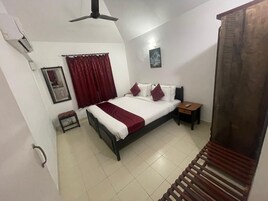 Quarto com cama, mesa de cabeceira, ventoinha no teto, janela com cortinas e um quadro na parede.