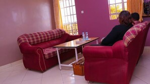 Quarto com sofá vermelho, uma mesa pequena, duas pessoas sentadas e uma janela com cortinas.