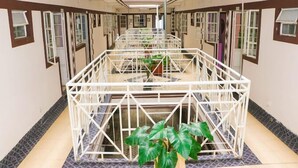 Um corredor com um caminho central, ladeado por janelas e portas, que leva a um quarto com cama e planta.