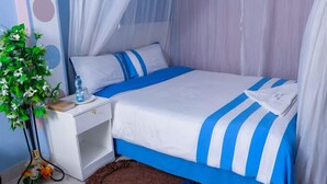 Uma cama bem arrumada com roupa de cama azul e branca, uma mesinha de cabeceira com uma garrafa de água e uma planta decorativa.