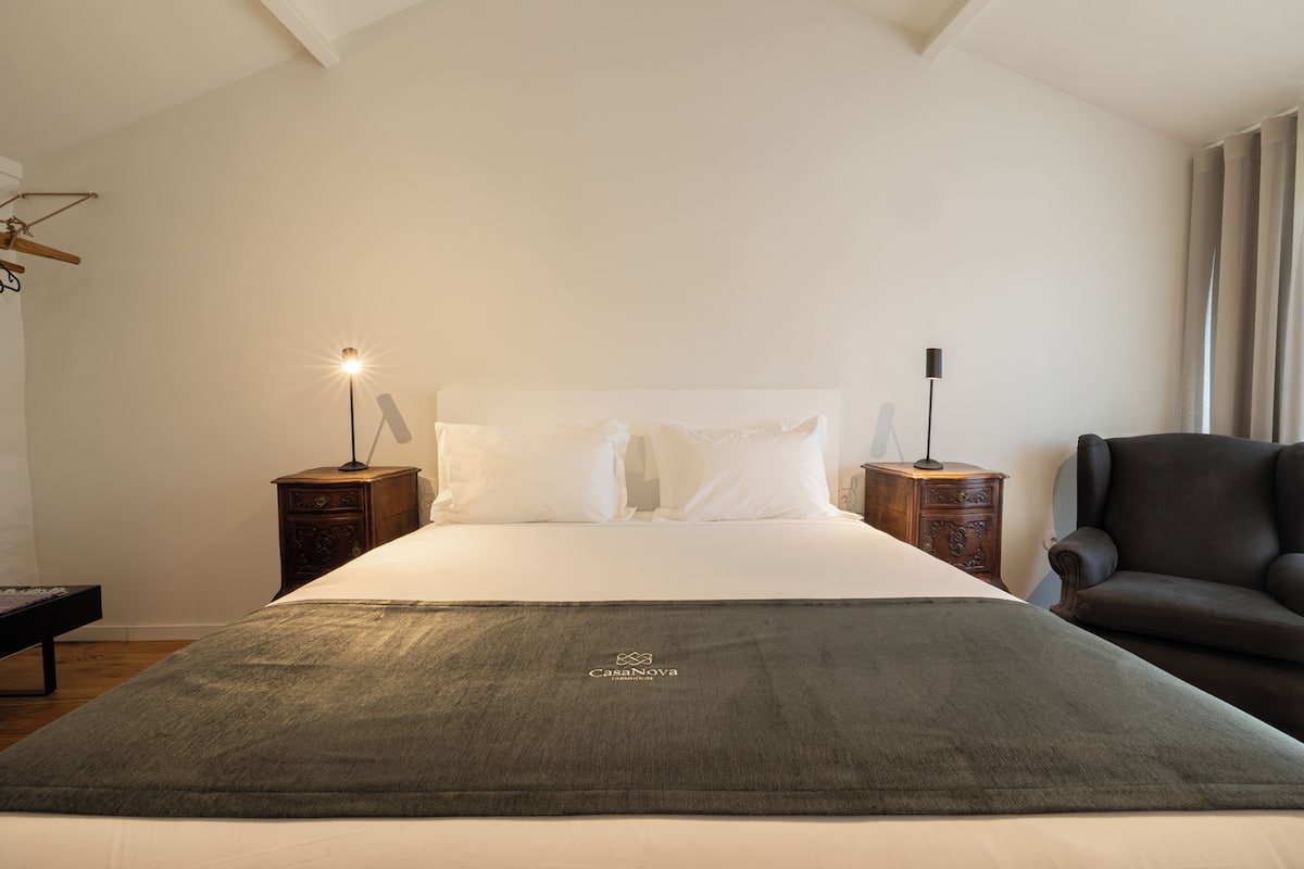 Cama bem arrumada com um cobertor cinzento, duas mesas de cabeceira com luminárias e uma poltrona escura num quarto moderno com paredes brancas e teto inclinado.