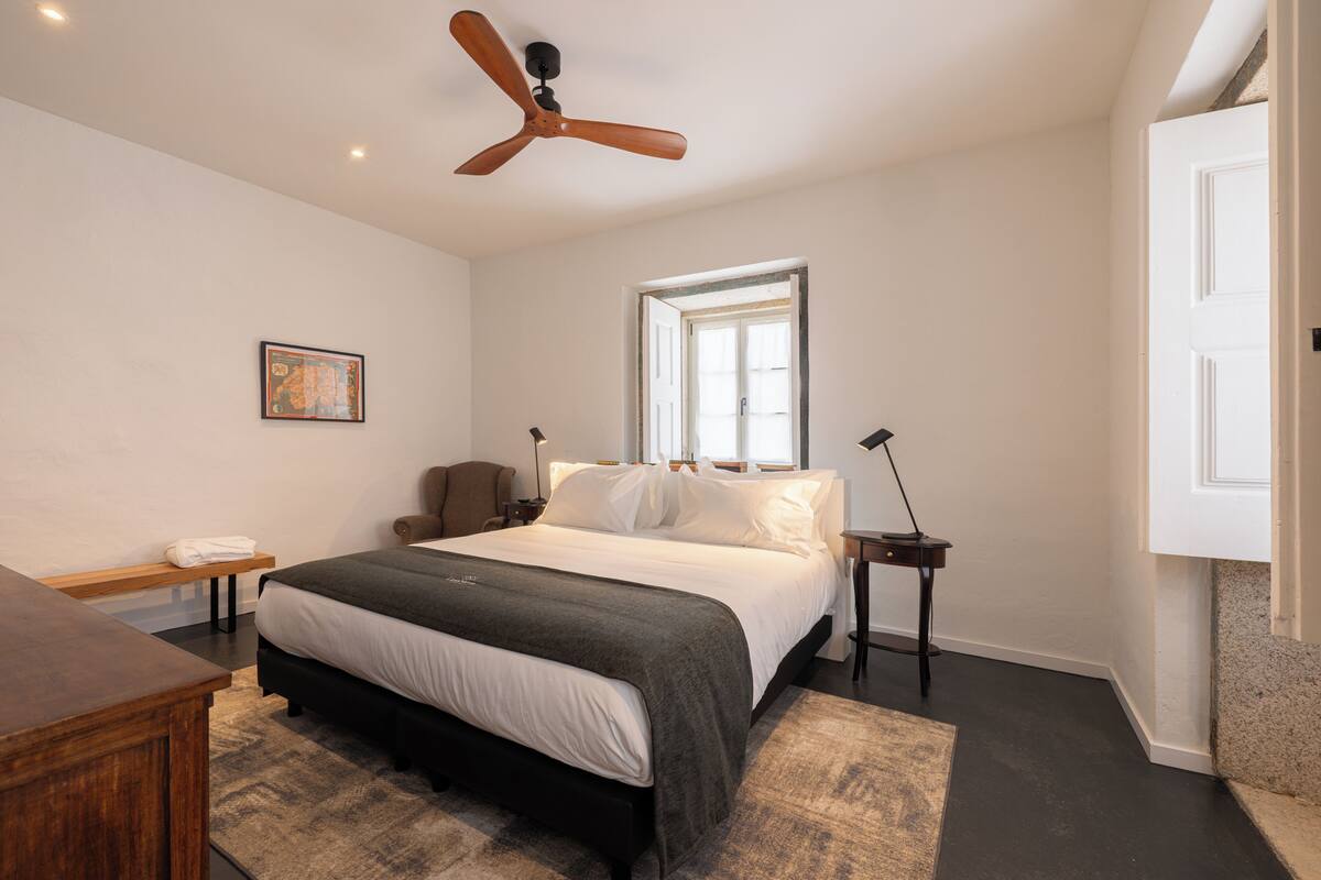Quarto com cama, escrivaninha, cadeira, ventoinha no teto, janela e porta.