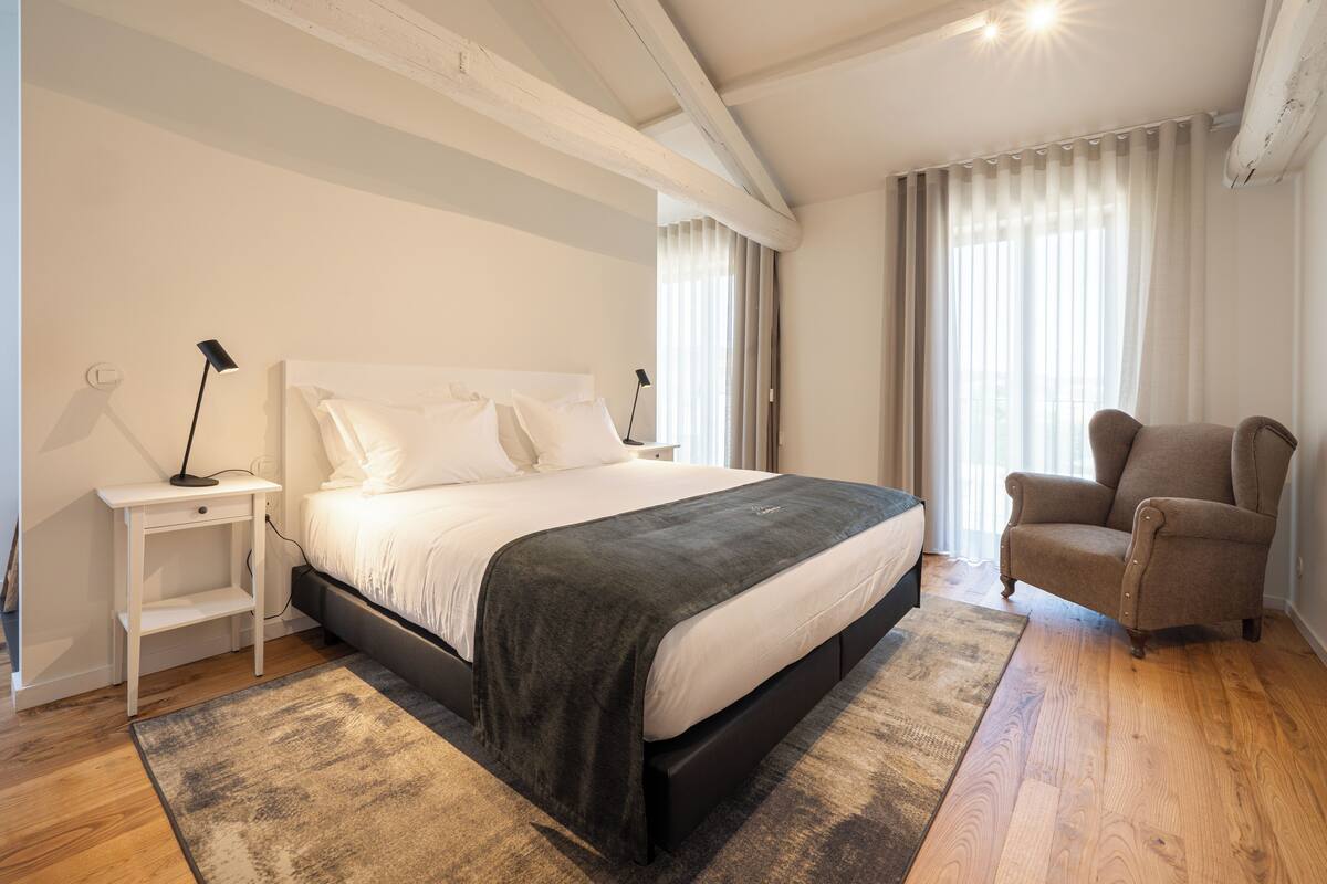 Quarto com cama, mesas de cabeceira, uma cadeira e janela com cortinas.
