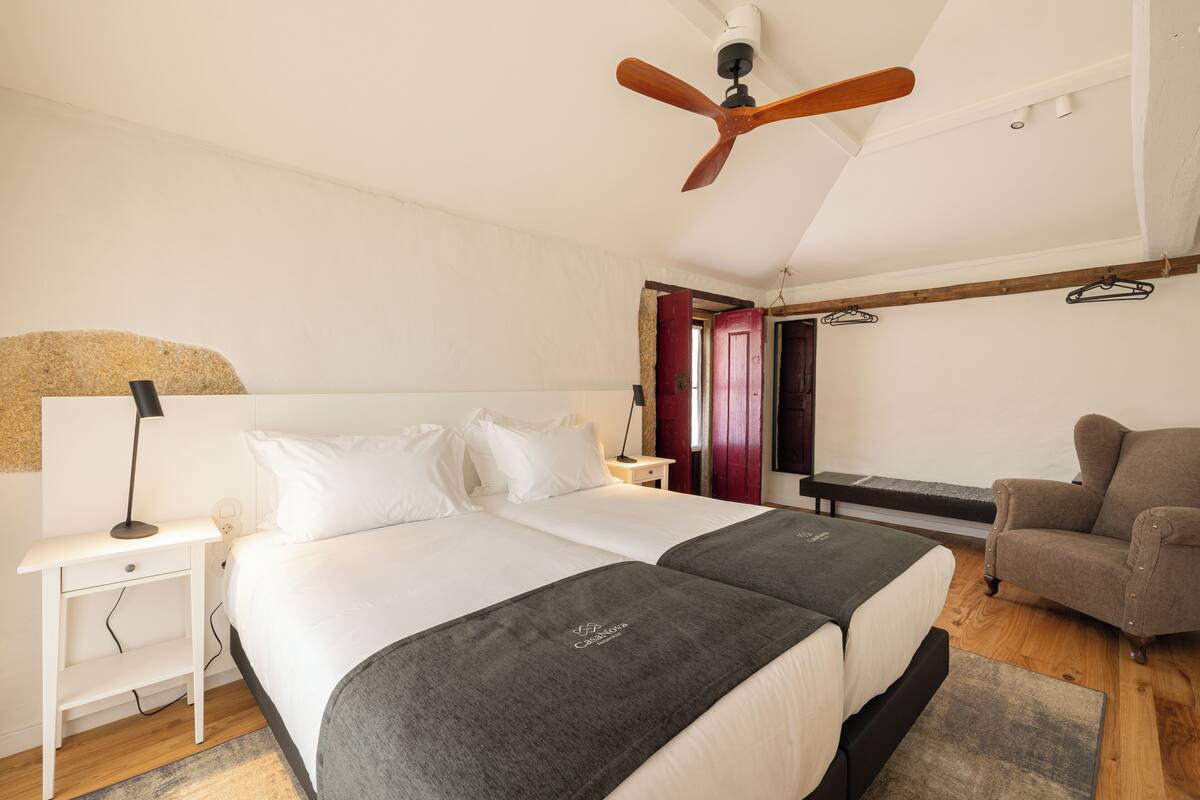 Quarto com duas camas, um ventilador de teto, mesa de cabeceira, uma cadeira e um banco de madeira.