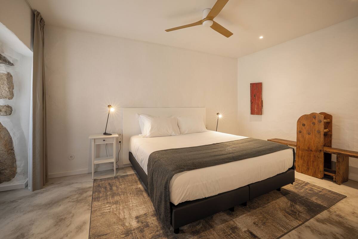 Quarto com uma cama grande, mesas de cabeceira, ventilador de teto e um banco de madeira.