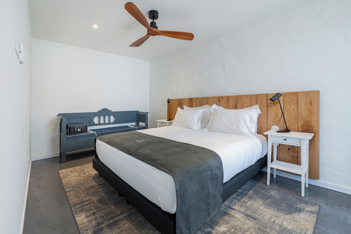 Quarto moderno com uma cama grande, cabeceira de madeira, mesa de cabeceira com abajur e ventilador de teto.