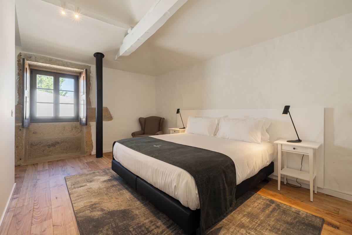 Um quarto com uma cama grande, uma mesinha de cabeceira, uma cadeira e uma janela com vista.