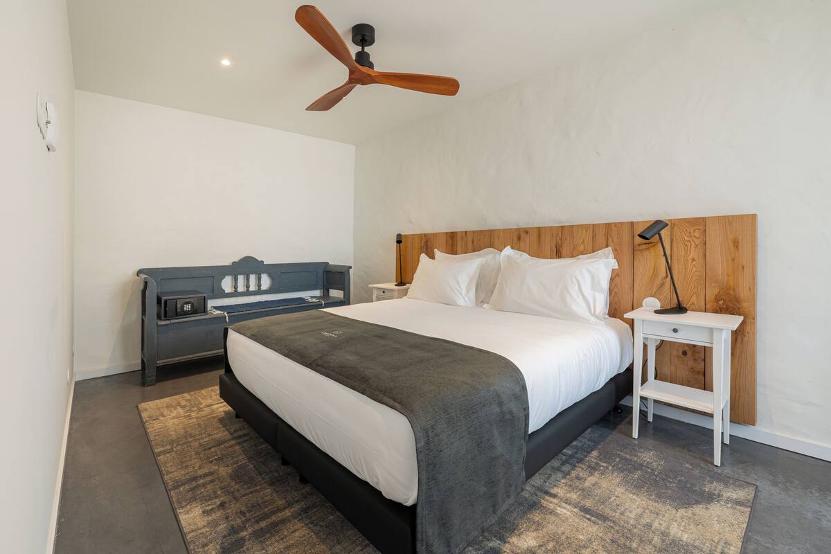 Quarto moderno com uma cama grande, cabeceira de madeira, mesa de cabeceira com abajur e ventoinha de teto.