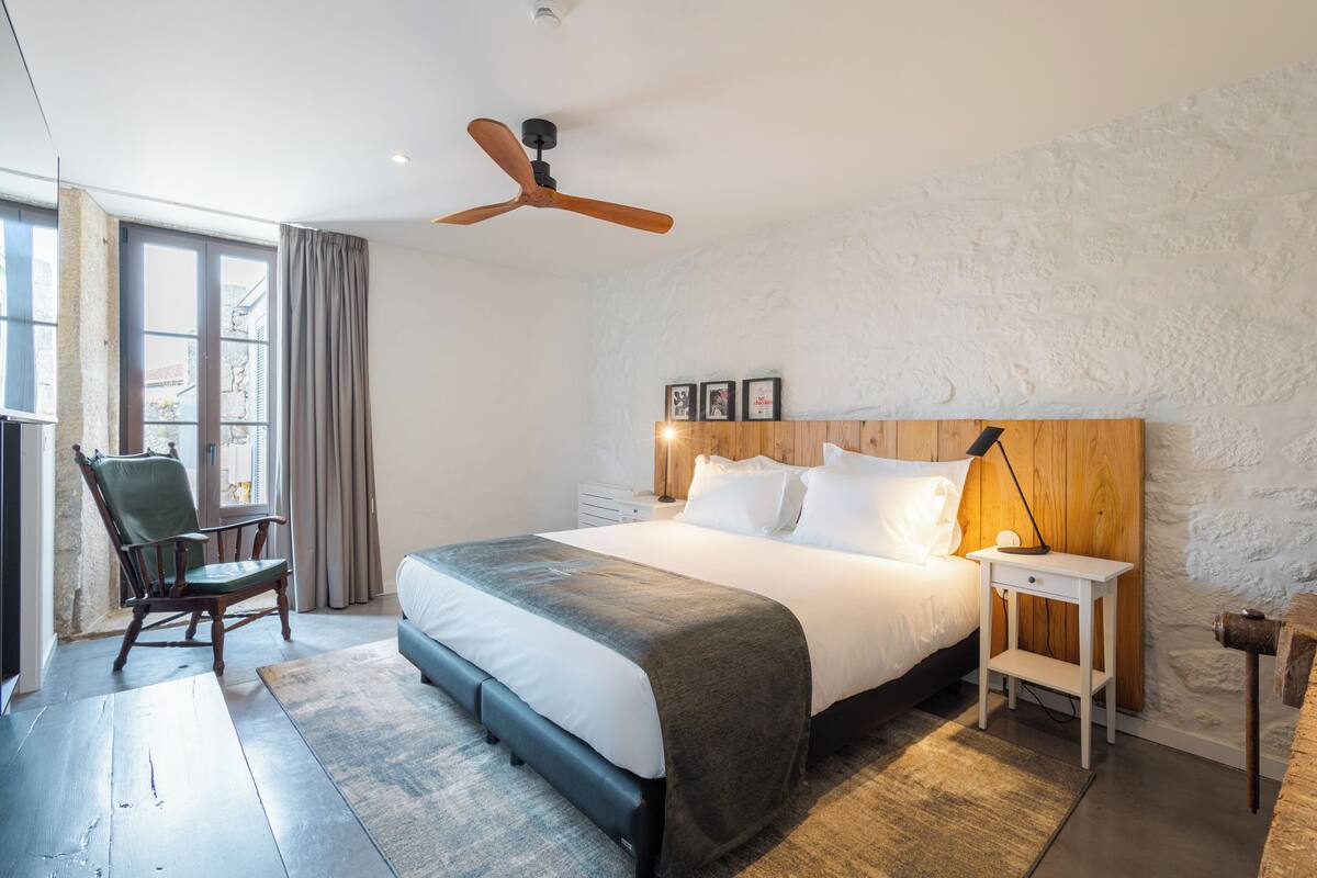 Quarto moderno com uma cama grande, cabeceira de madeira, ventilador de teto, uma cadeira e uma mesinha de cabeceira.