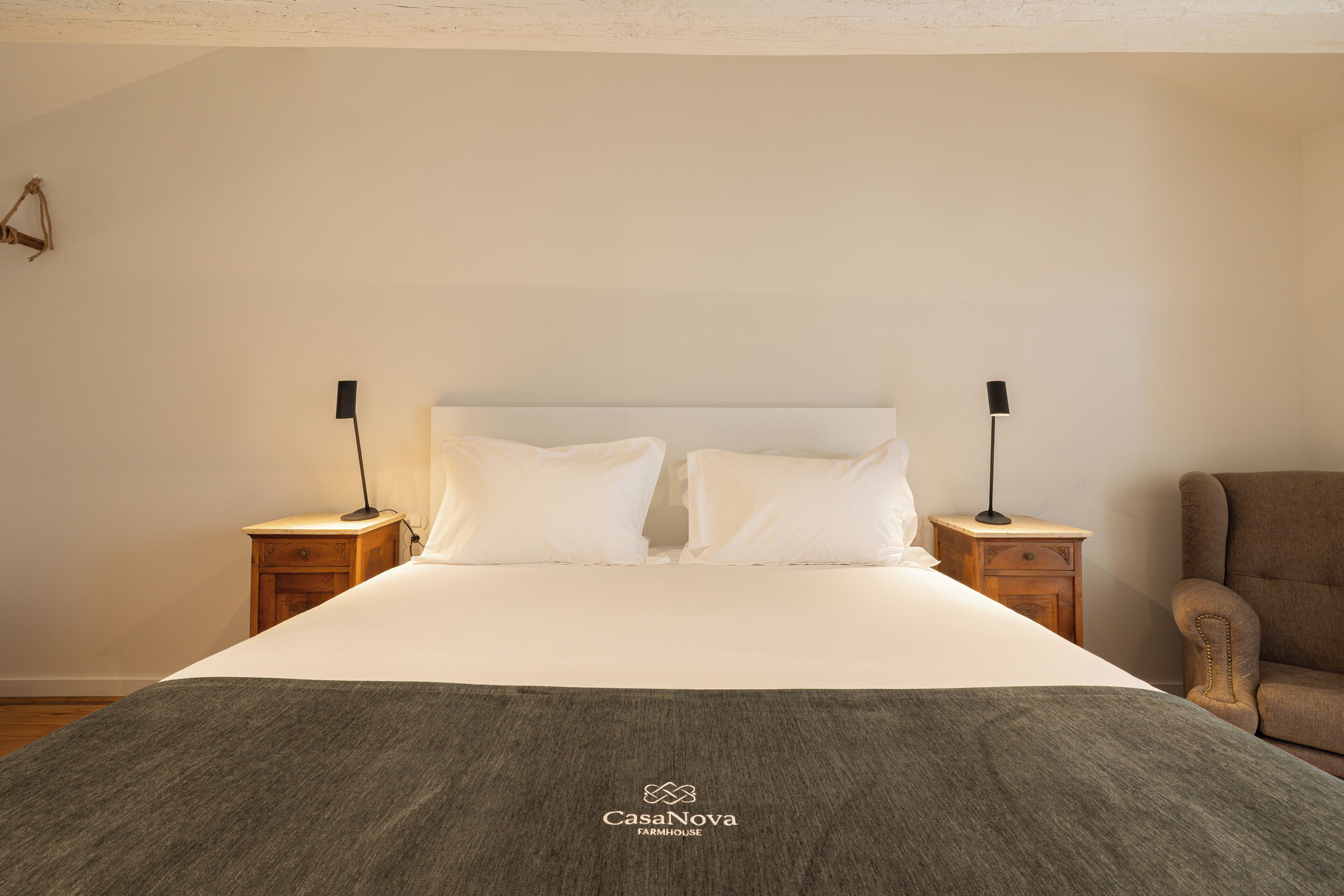 Cama bem arrumada com lençóis brancos e manta cinza, acompanhada por duas mesas de cabeceira com abajures, em um quarto com parede branca lisa e piso de madeira.