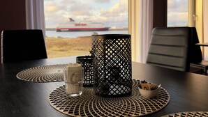 Uma mesa com uma garrafa, um copo e um castiçal, com vista para um navio e um céu nublado através de uma janela.