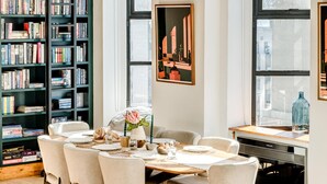 Uma área de refeições com uma mesa de madeira posta para quatro pessoas, rodeada por cadeiras, com uma estante de livros cheia de livros à esquerda e uma obra de arte emoldurada acima da mesa.