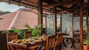 Uma área de refeições ao ar livre com mesas e cadeiras de madeira, cercada por vasos de plantas e um painel decorativo.