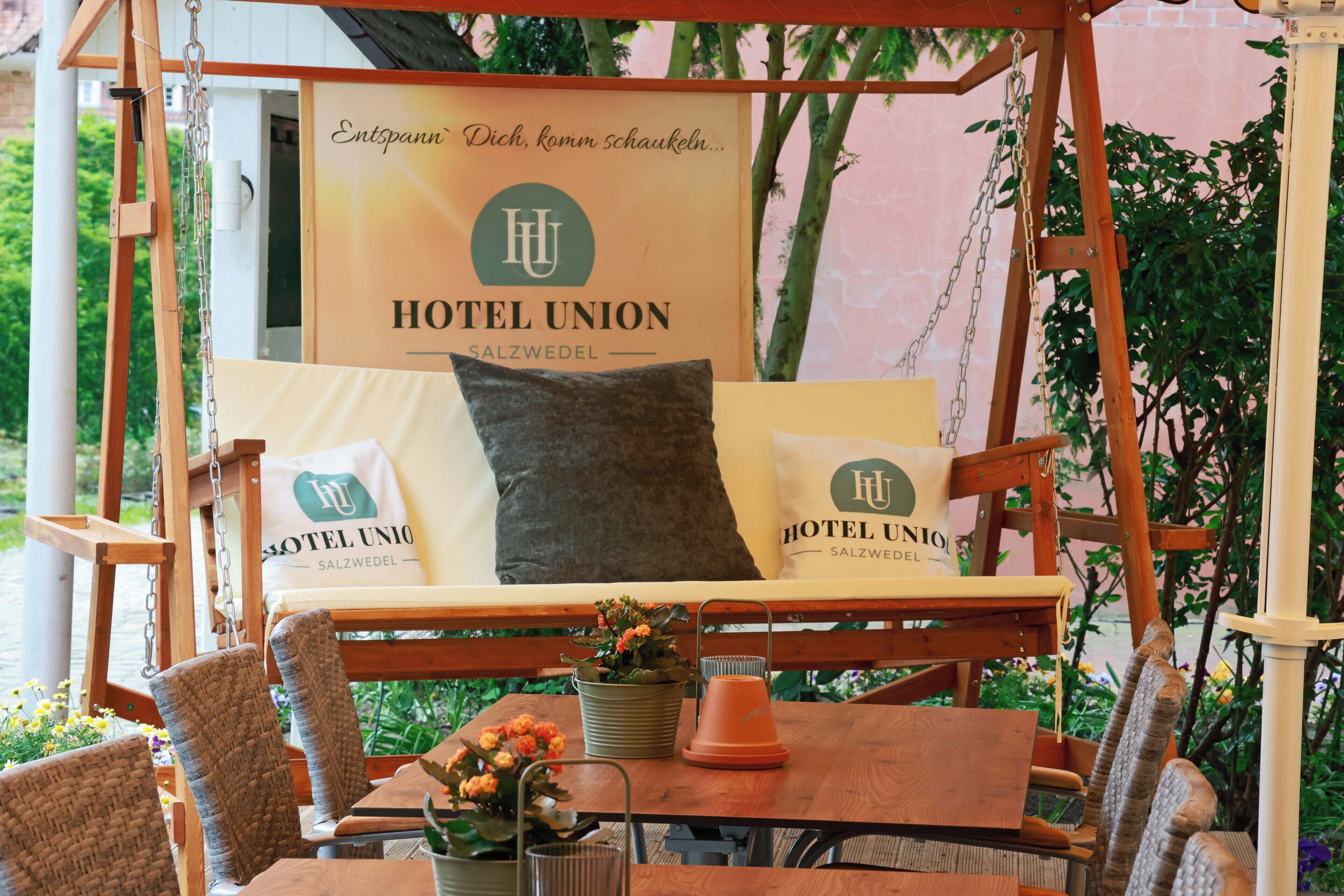 Uma cadeira balanço de madeira com almofada, uma mesa de metal com planta em vaso, e um banner do Hotel Union Salzburgwedel.