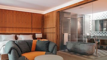 Quarto de hotel moderno com cabeceira de madeira, uma cama grande, área de estar com um banco e almofadas, banheira de pedra e um espelho grande sobre a pia.