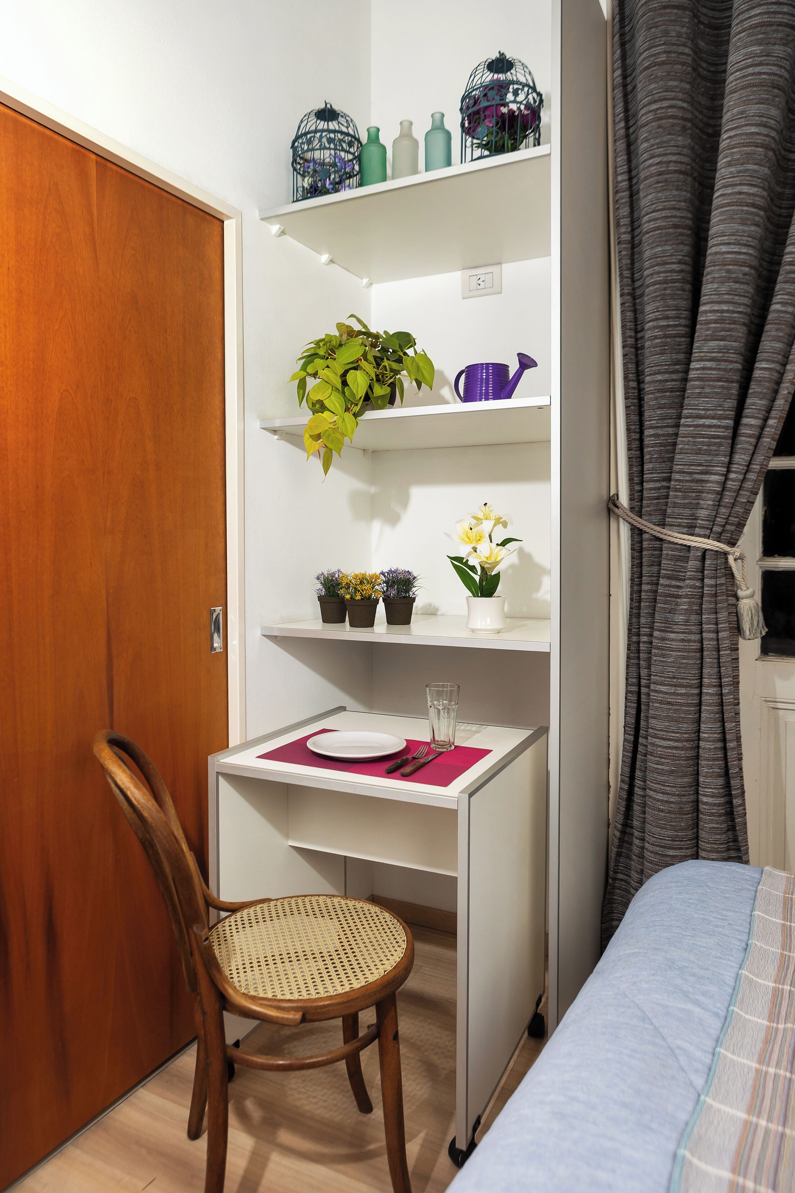 Quarto compacto com porta de madeira, escrivaninha branca com cadeira, cama com cobertor listrado e prateleira com objetos decorativos.
