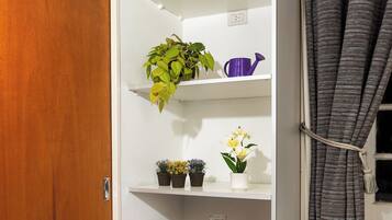 Quarto compacto com porta de madeira, escrivaninha branca com cadeira, cama com cobertor listrado e prateleira com objetos decorativos.