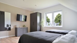 Quarto moderno com duas camas, televisão, guarda-roupa e janela com vista para árvores.