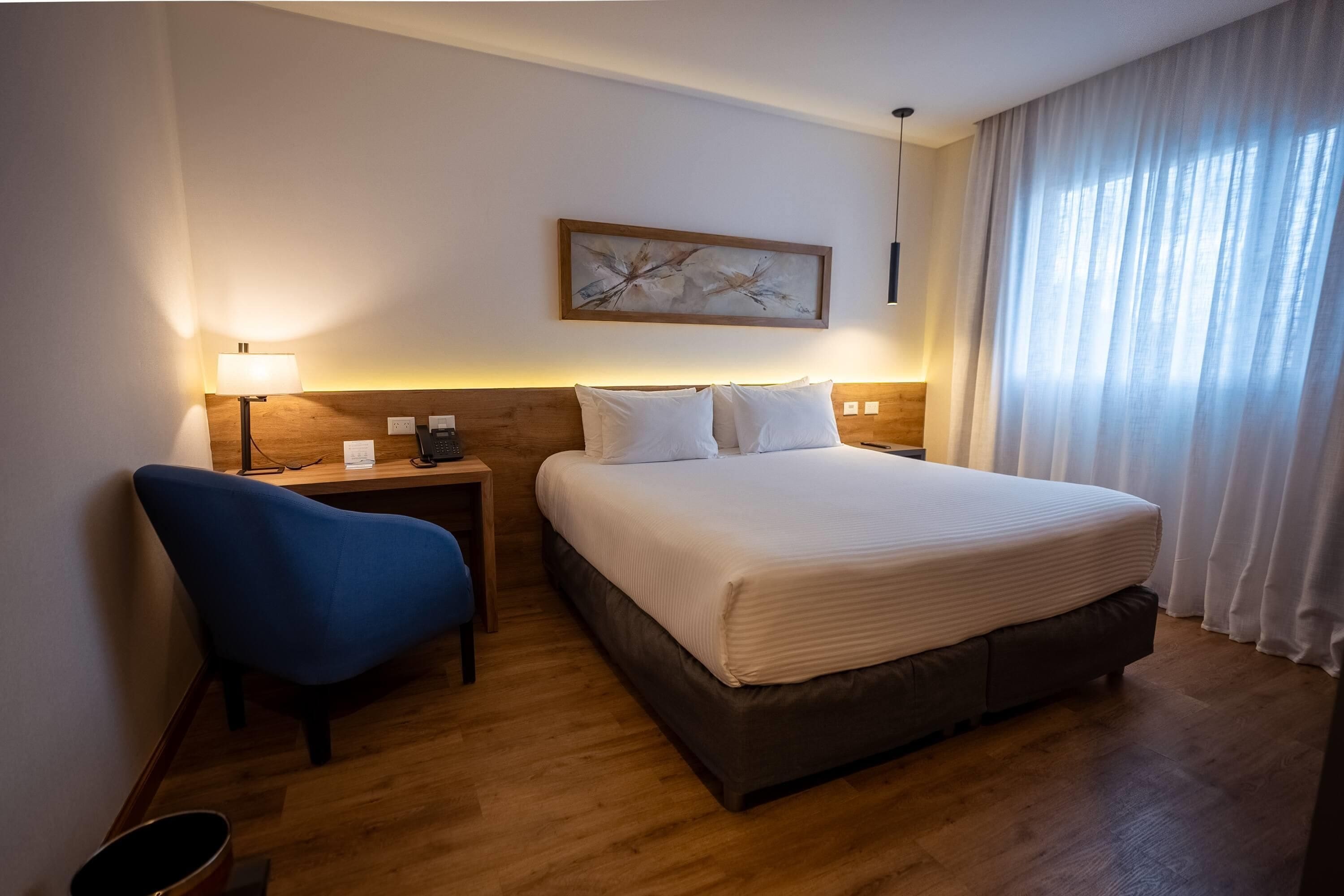 Quarto de hotel com cama, escrivaninha, cadeira, abajur e um quadro na parede.