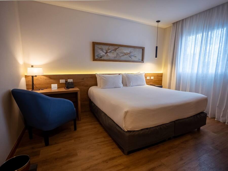 Quarto de hotel com cama, escrivaninha, cadeira, abajur e um quadro na parede.