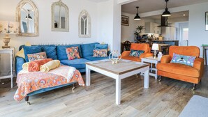 Uma sala de estar com um sofá azul, uma poltrona laranja, uma mesa de centro de madeira e uma cozinha ao fundo.