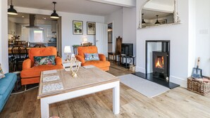 Uma sala de estar com dois sofás laranja, um sofá azul, uma mesa de centro de madeira, uma lareira e uma cozinha ao fundo.