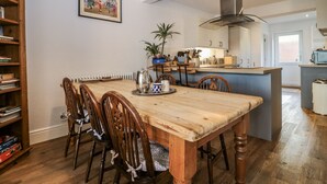 Uma área de refeições com uma mesa e cadeiras de madeira, uma estante de livros, um vaso com planta e uma cozinha ao fundo.