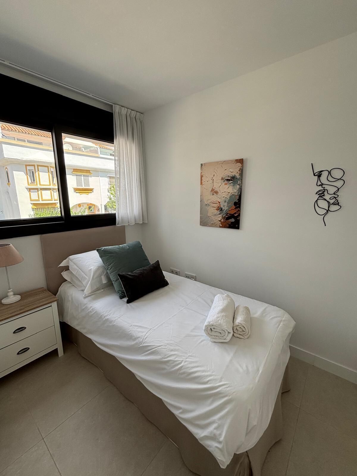 Cama bem arrumada com lençóis brancos, um travesseiro e toalhas dobradas. Há uma mesa de cabeceira com uma luminária e uma obra de arte na parede.