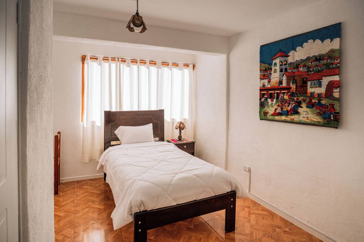 Quarto com cama de madeira, roupa de cama branca, mesa de cabeceira com abajur e um quadro na parede.
